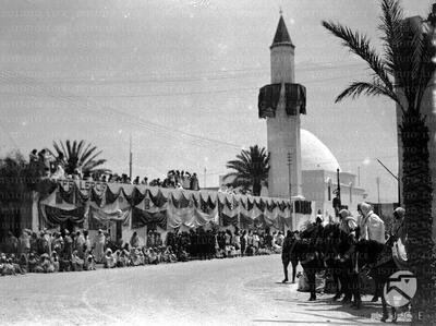 Homs (?) crowd of indigenous people waits along a street, with minaret ...
